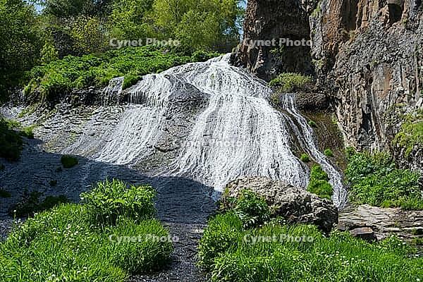 A bright waterfall snakes through green plants and rocks in sunny ...